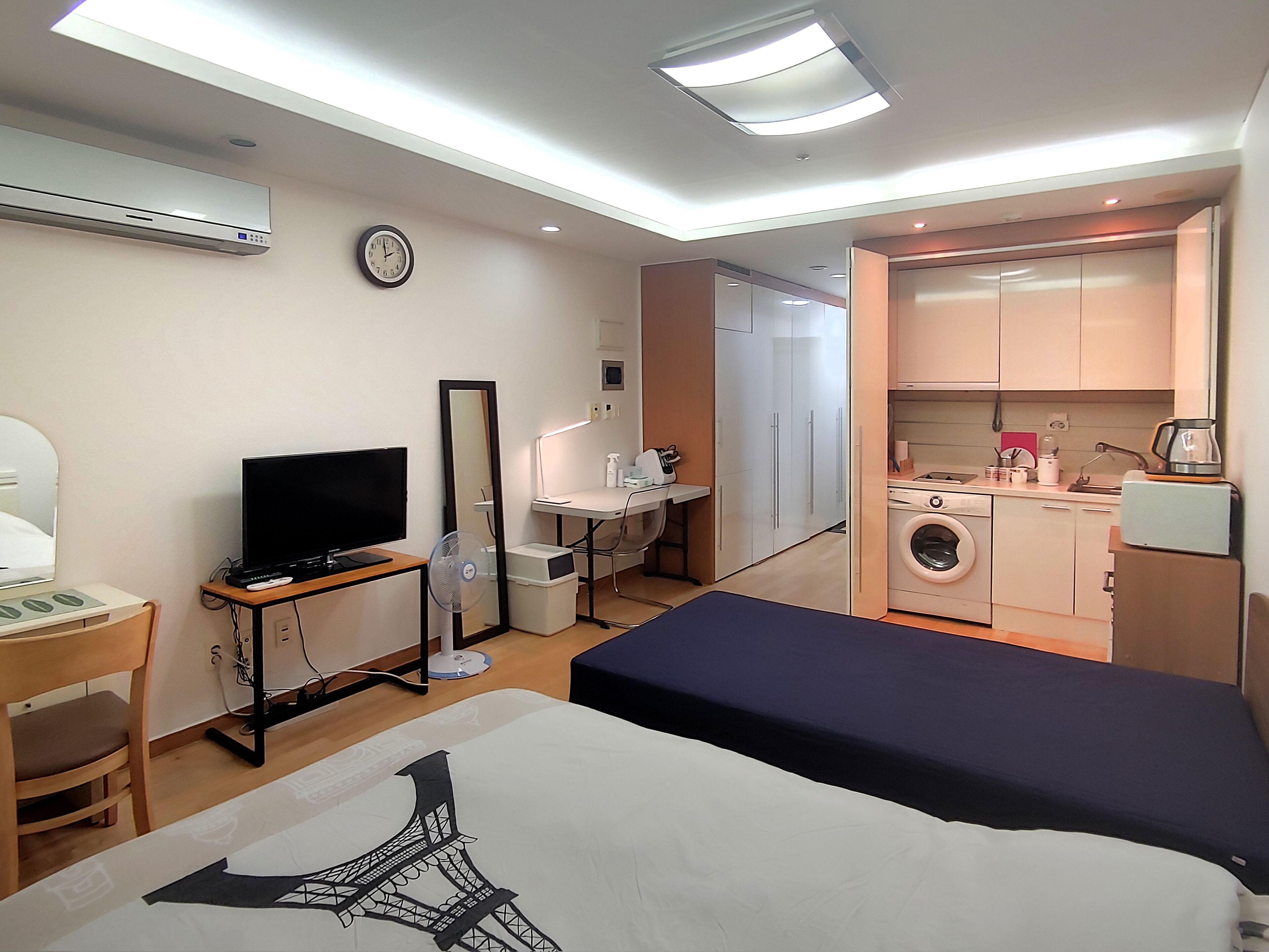 Quarto de hotel com cama, escrivaninha com televisão, espelho, ventoinha, máquina de lavar e cozinha compacta.