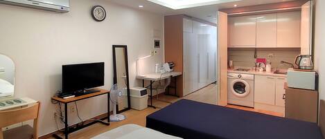 Quarto de hotel com cama, escrivaninha com televisão, espelho, ventoinha, máquina de lavar e cozinha compacta.