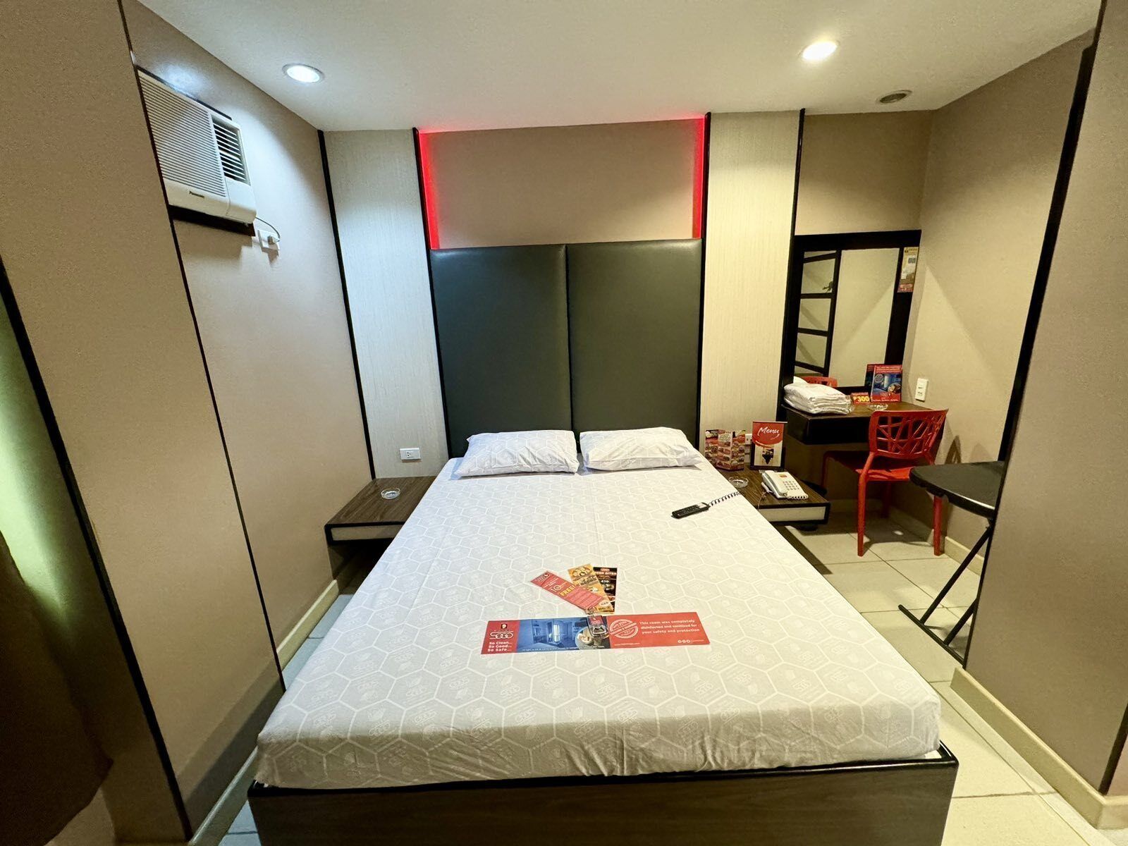 Quarto de hotel com cama, escrivaninha, cadeira, espelho e mesa de cabeceira.