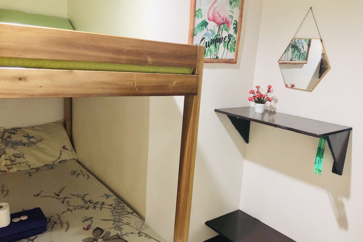 Uma cama beliche com colcha estampada, uma pequena prateleira com espelho e um vaso de planta, e um porta-retrato fixado na parede.
