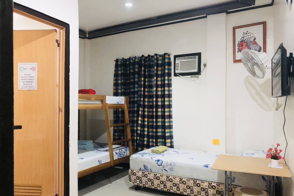 Quarto com uma cama de solteiro, uma escrivaninha, uma cadeira, um banquinho pequeno, um ventilador e um quadro na parede.