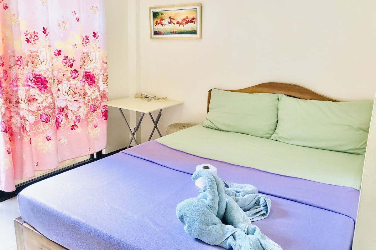 Quarto com cama, uma mesinha, uma cadeira e cortina estampada com motivos florais.
