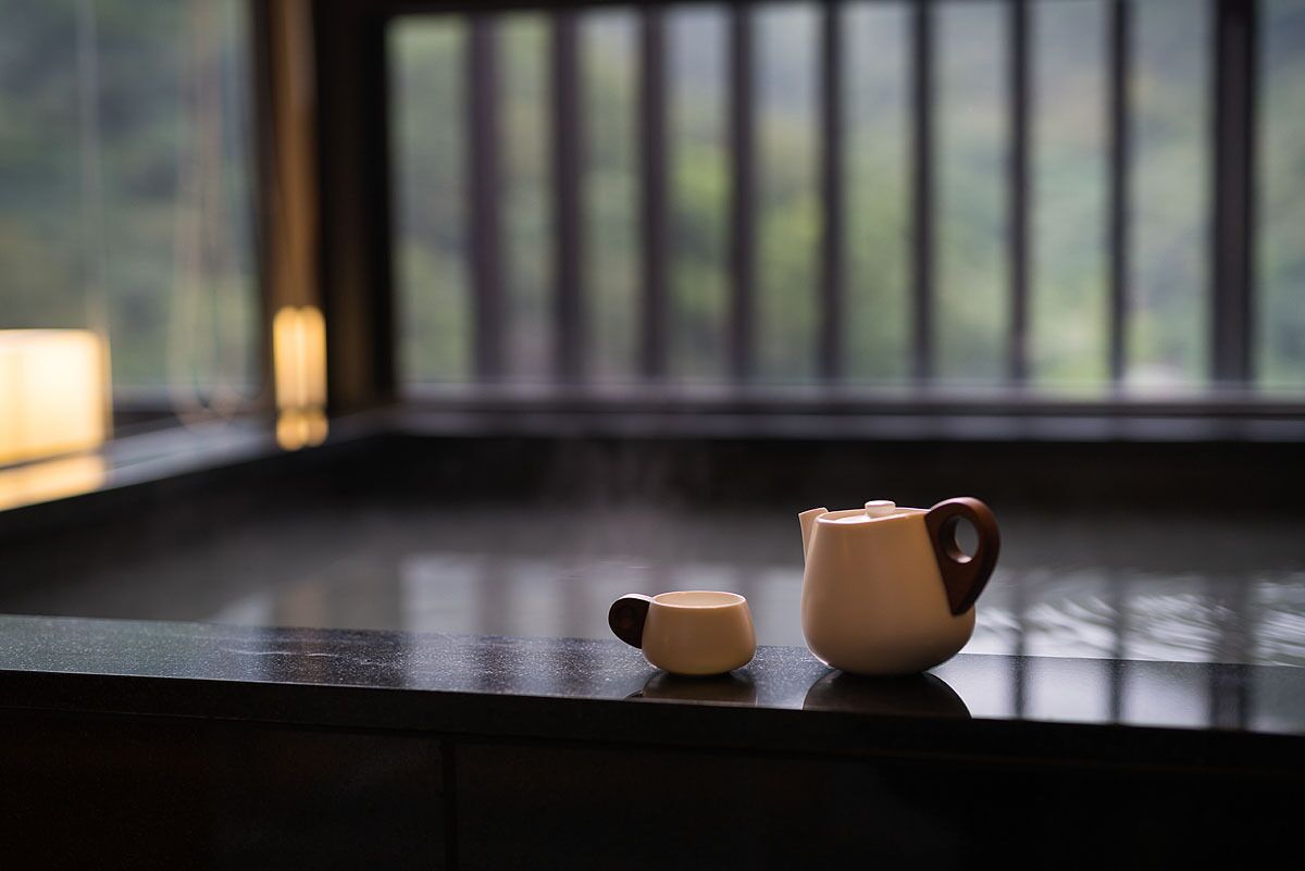 Uma mesa com um bule e uma chávena, colocada perto de uma janela com vista para árvores.