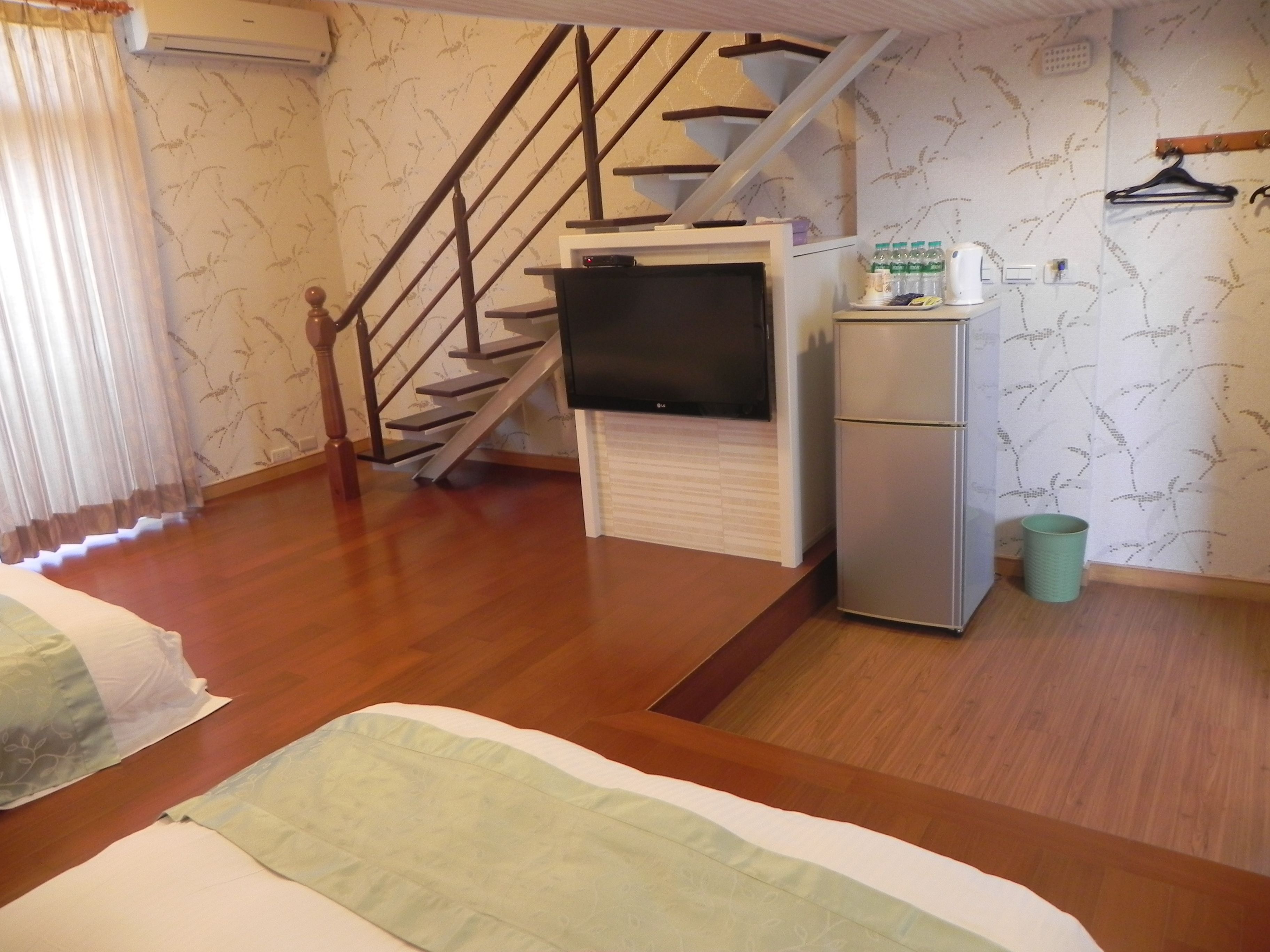 Quarto de hotel com piso de madeira, cama com roupa de cama verde, televisão, mini-geladeira, escada e ar-condicionado fixado na parede.