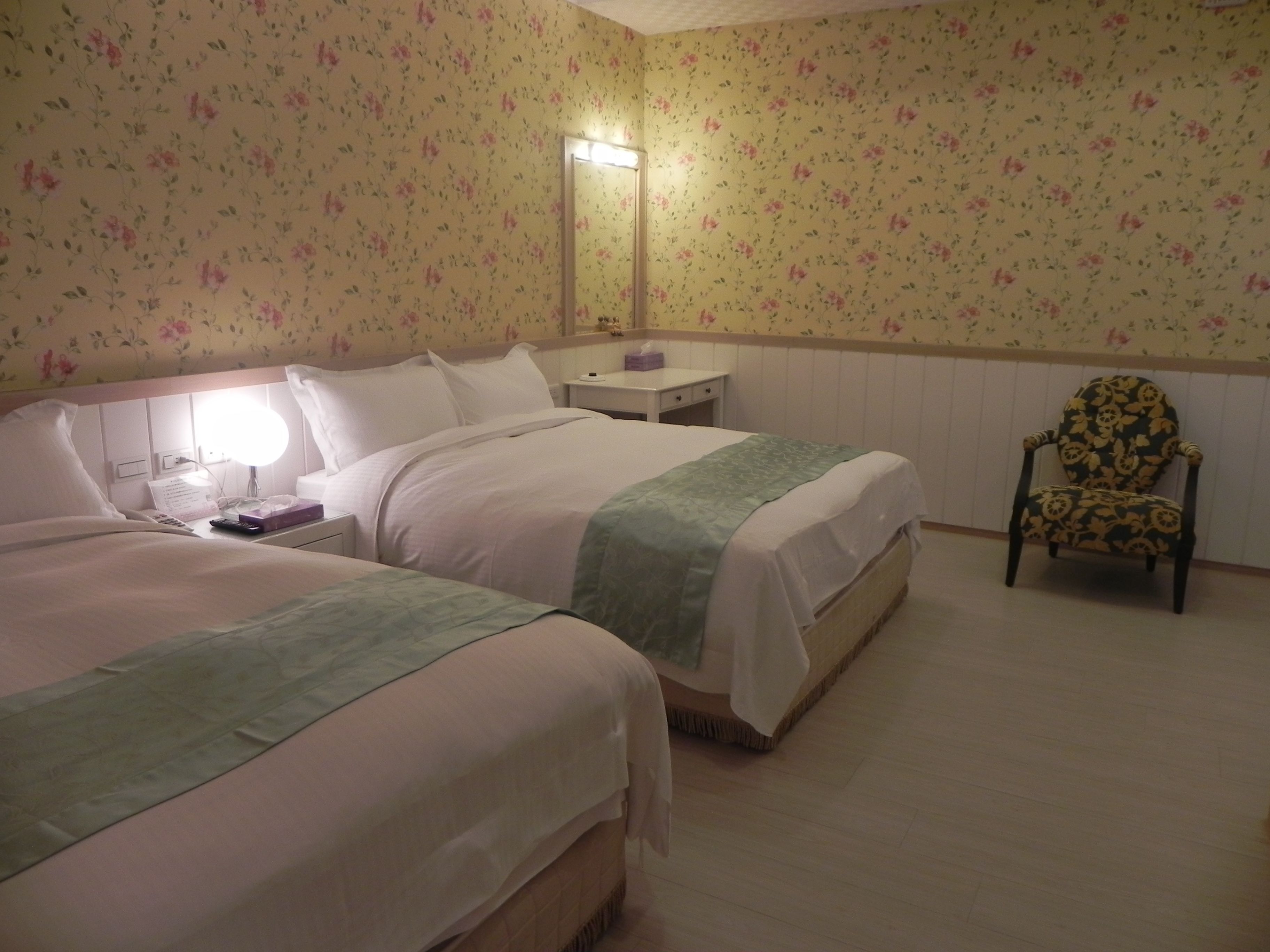 Quarto de hotel com duas camas, uma mesinha de cabeceira, uma cadeira e papel de parede floral.