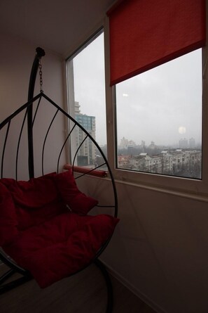 Quarto com uma cadeira suspensa, uma janela grande com vista para a paisagem urbana e uma persiana vermelha.