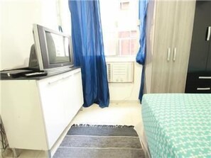 Um quarto pequeno com uma cama, uma escrivaninha com um computador e uma janela com cortinas azuis.