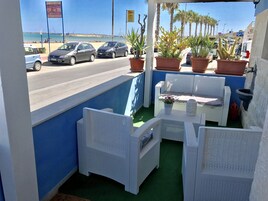 Uma varanda com móveis de vime branco, plantas em vasos e vista para a praia e carros na rua.