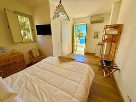 Quarto com cama, televisão, cadeira de vime, penteadeira de madeira e porta que dá para uma varanda.