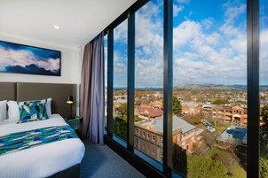 Quarto de hotel com uma cama grande, vista da cidade através de janelas do chão ao teto e um quadro na parede.
