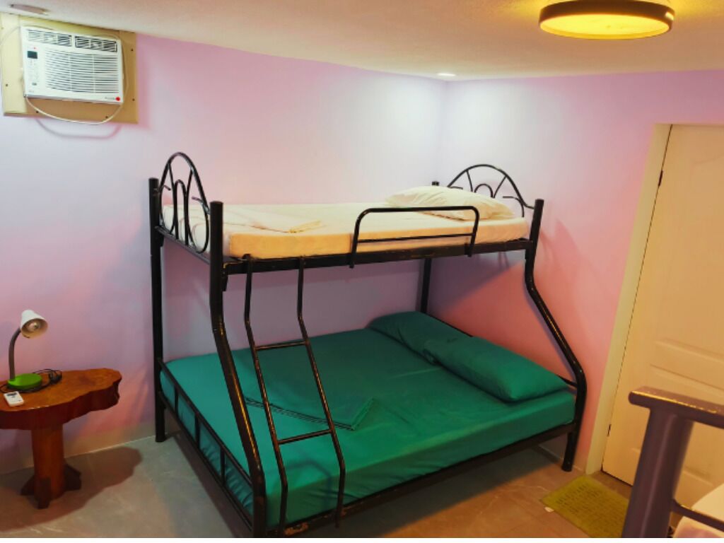 Uma cama beliche com colchão verde, uma mesinha com luminária e um ar condicionado fixado na parede.