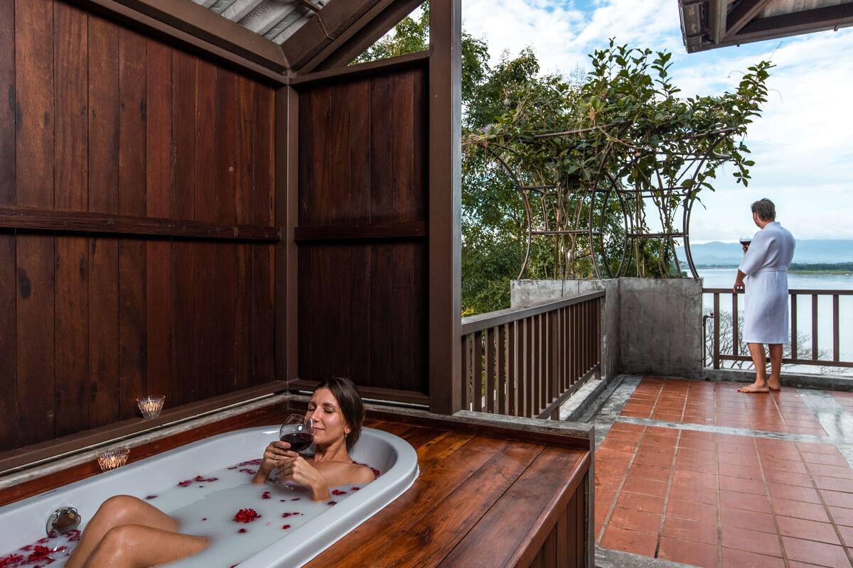 Mulher relaxando em banheira cheia de água e pétalas de rosa, com um homem em varanda apreciando uma vista panorâmica.