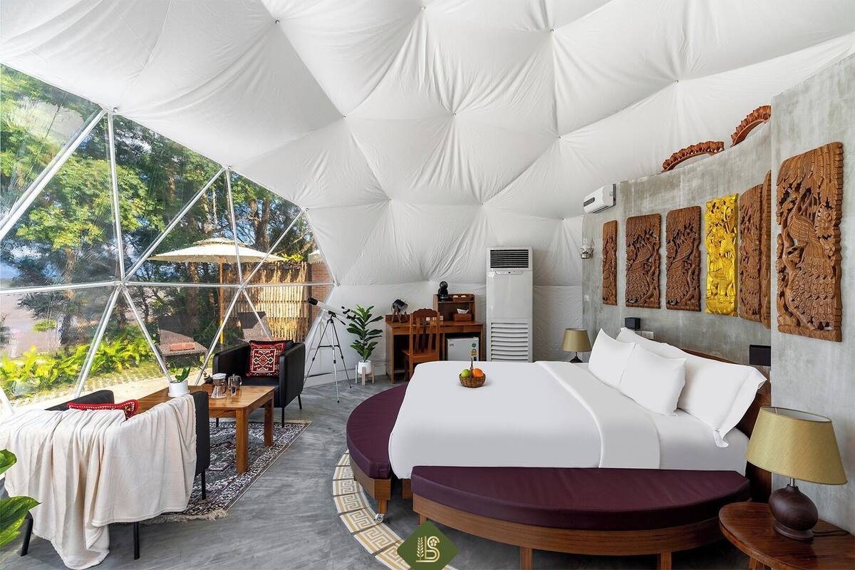 Um quarto espaçoso coberto por uma tenda, com uma cama, uma escrivaninha e vista para o verde lá fora.