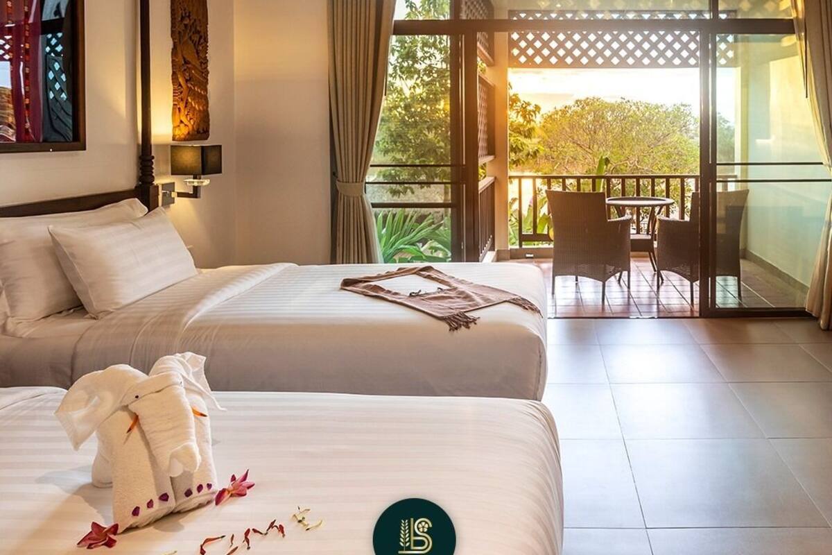 Quarto de hotel com duas camas, varanda com mesa e cadeiras, e vista para o verde.