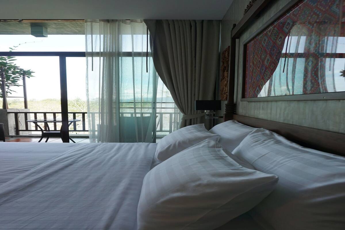 Quarto de hotel com uma cama grande, varanda com mesa e cadeiras, e vista para o exterior.