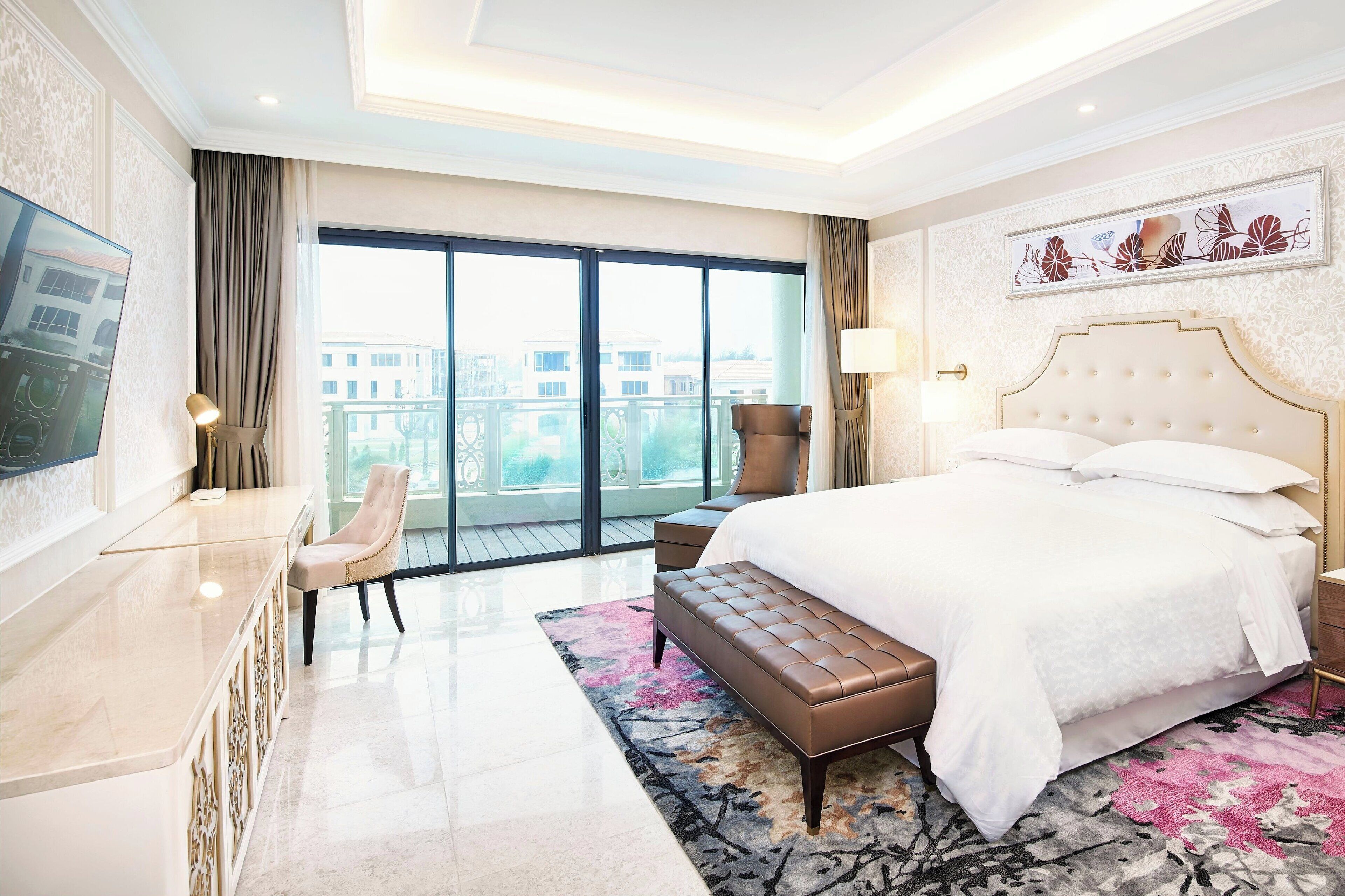 Quarto de hotel com uma cama grande, uma escrivaninha, uma cadeira, um banco, uma TV e varanda com vista para prédios.