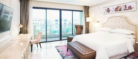 Quarto de hotel com uma cama grande, uma escrivaninha, uma cadeira, um banco, uma TV e varanda com vista para prédios.