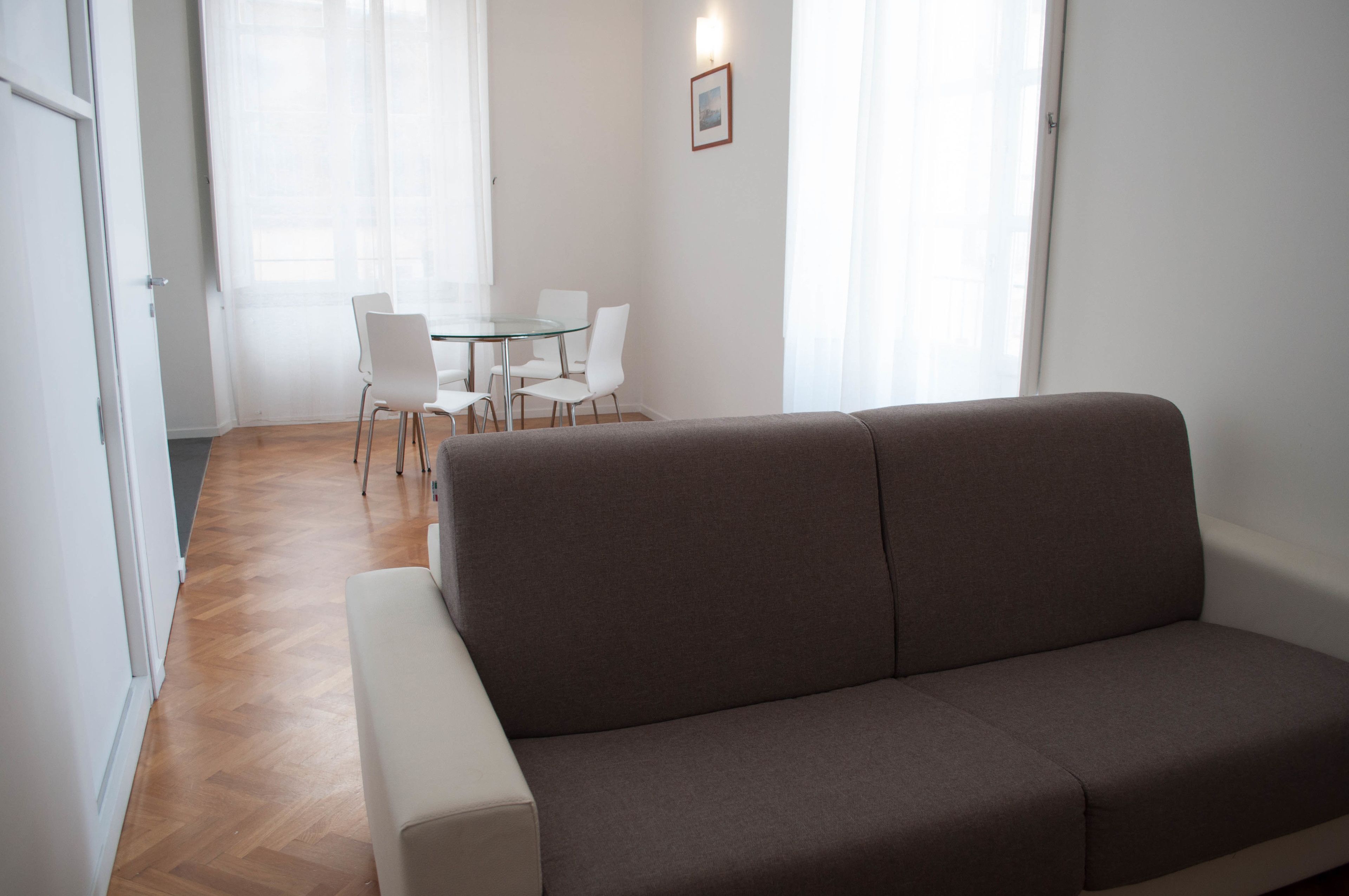 Uma sala de estar com um sofá marrom, uma mesa de vidro com cadeiras e uma parede branca com um quadro.