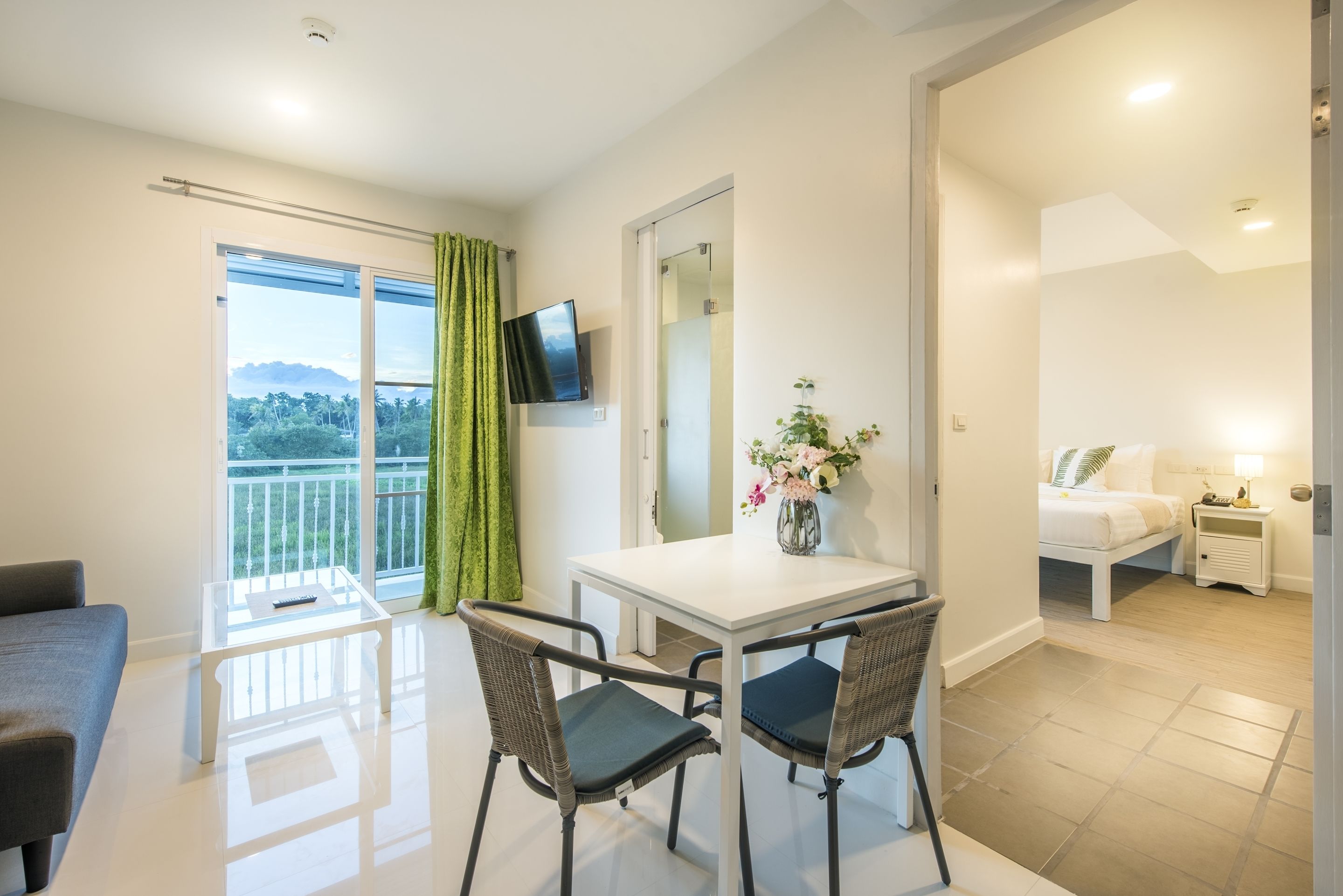Quarto de hotel moderno com varanda, área de refeições com mesa e cadeiras, cama com mesa de cabeceira e televisão na parede.