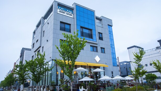 Um edifício moderno com fachada azul e cinza, um letreiro com 'RainBoots Guest House', área de estar externa com guarda-sóis e uma pequena árvore na frente.