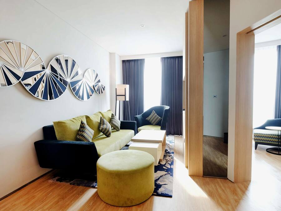 Uma sala de estar moderna com sofá, banqueta e mesa de centro. Há obras de arte abstratas nas paredes e vista para o exterior através de grandes janelas.