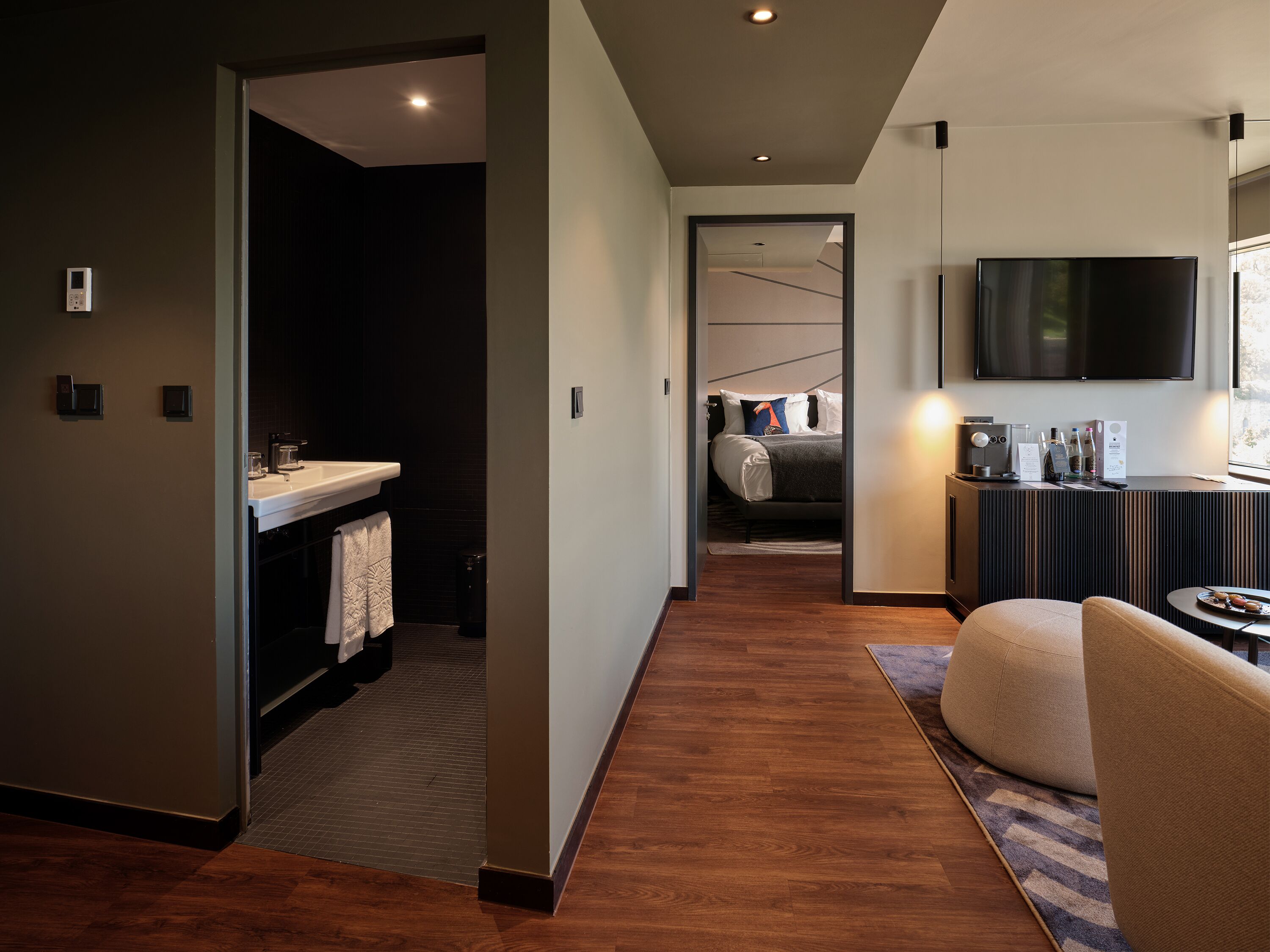 Quarto de hotel moderno com piso de madeira, uma cama grande, televisão e minibar.