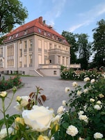 Um edifício grande e histórico com telhado vermelho e fachada simétrica, cercado por jardins exuberantes e rosas em flor.