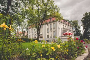 Um edifício histórico com telhado vermelho, rodeado por um jardim bem cuidado com diversas flores e um caminho de pedra.