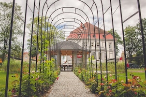 Um caminho no jardim com um arco de metal que leva a um edifício com telhado vermelho.