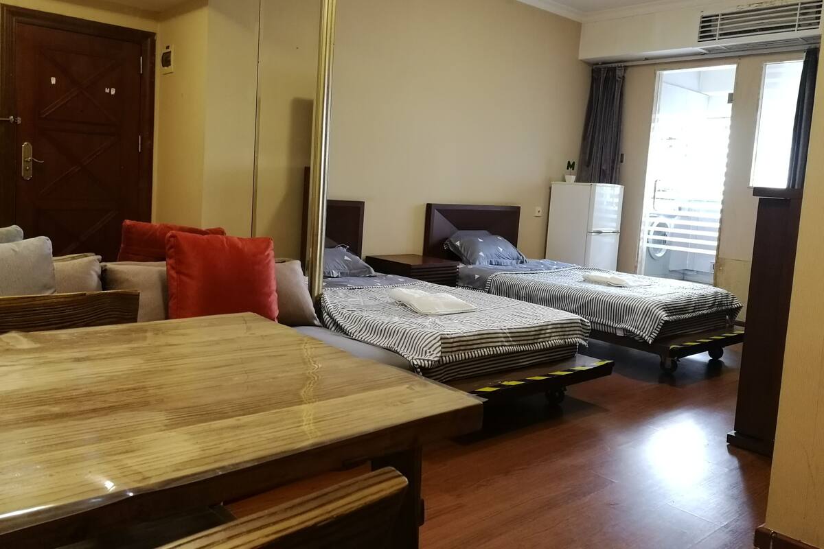 Quarto de hotel com duas camas, uma mesa de madeira, um espelho, um guarda-roupa e uma janela com persianas.