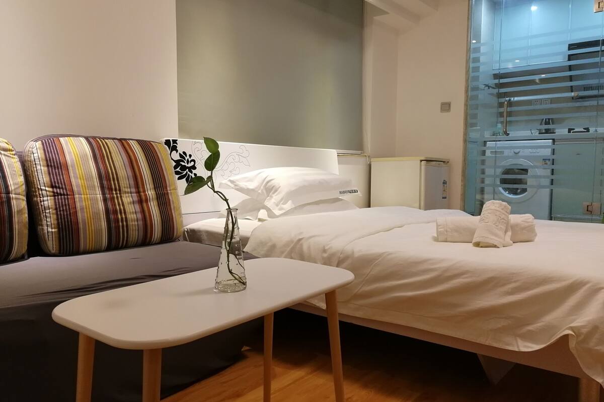 Quarto de hotel moderno com cama, uma mesinha, um sofá e área de lavanderia com paredes de vidro.