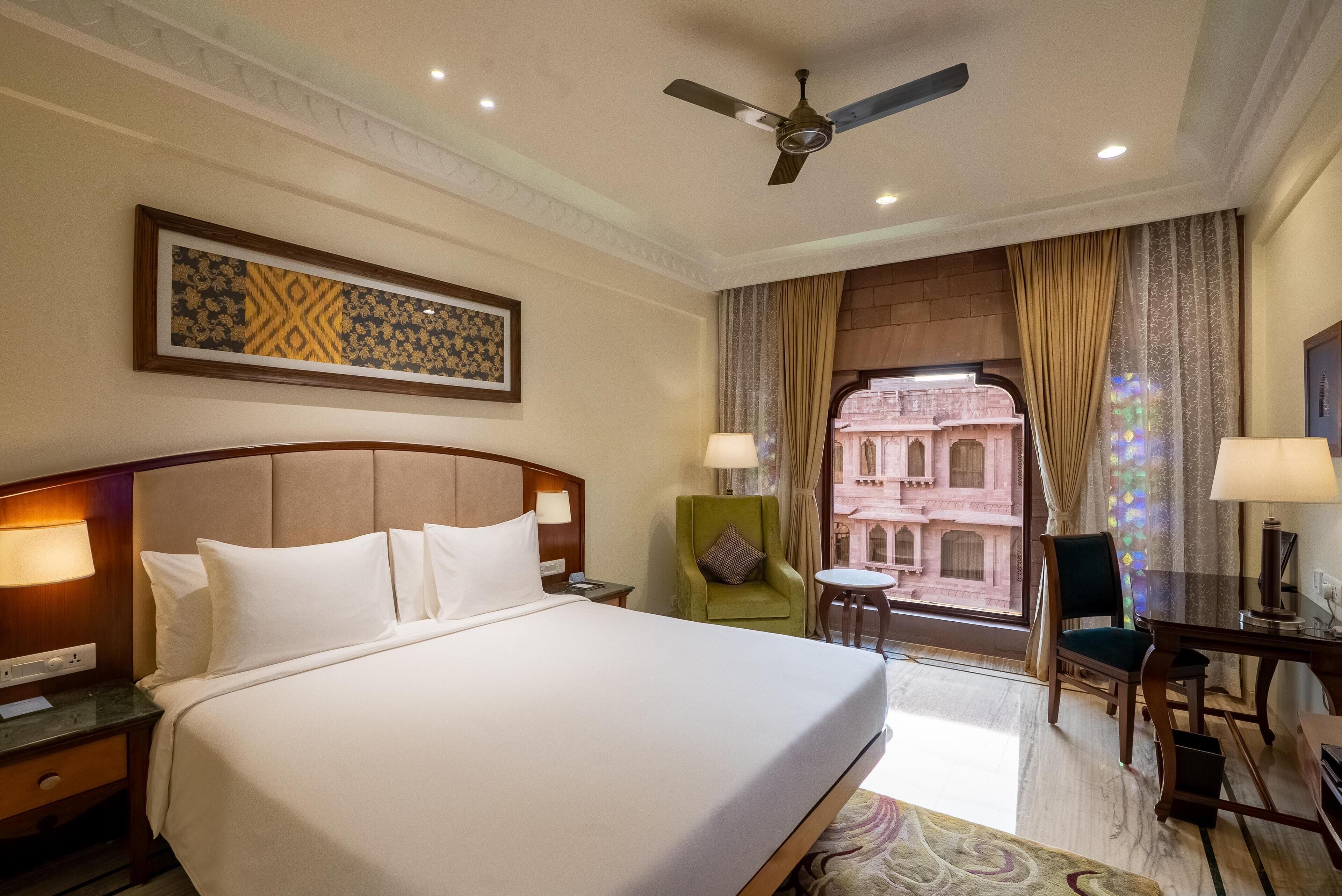 Quarto de hotel com uma cama grande, uma escrivaninha, uma cadeira, um ventilador de teto e vista para um edifício histórico.