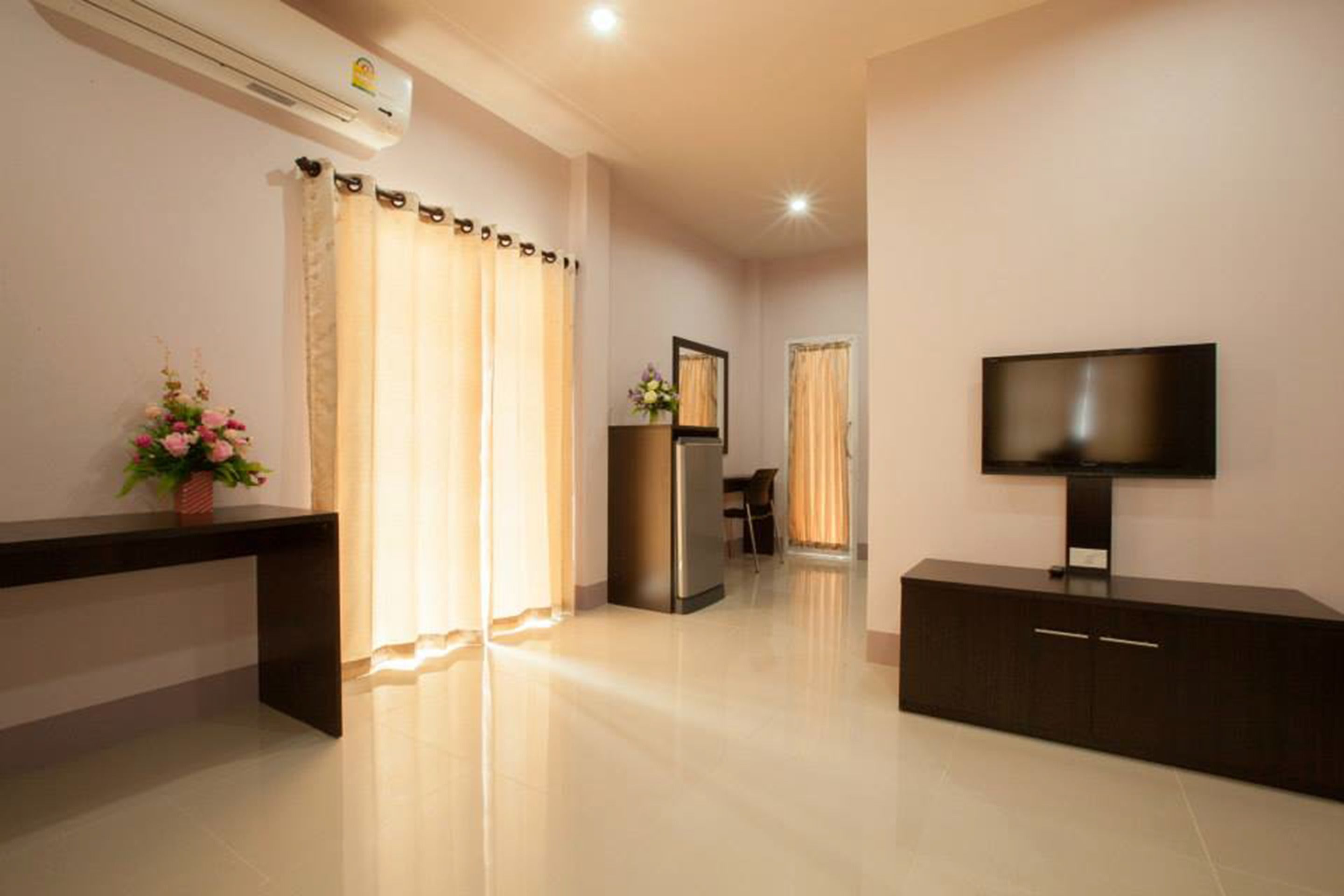 Uma sala de estar moderna com televisão de tela plana, um armário de madeira escura, uma mesa pequena com um vaso de flores e uma janela grande com cortinas.