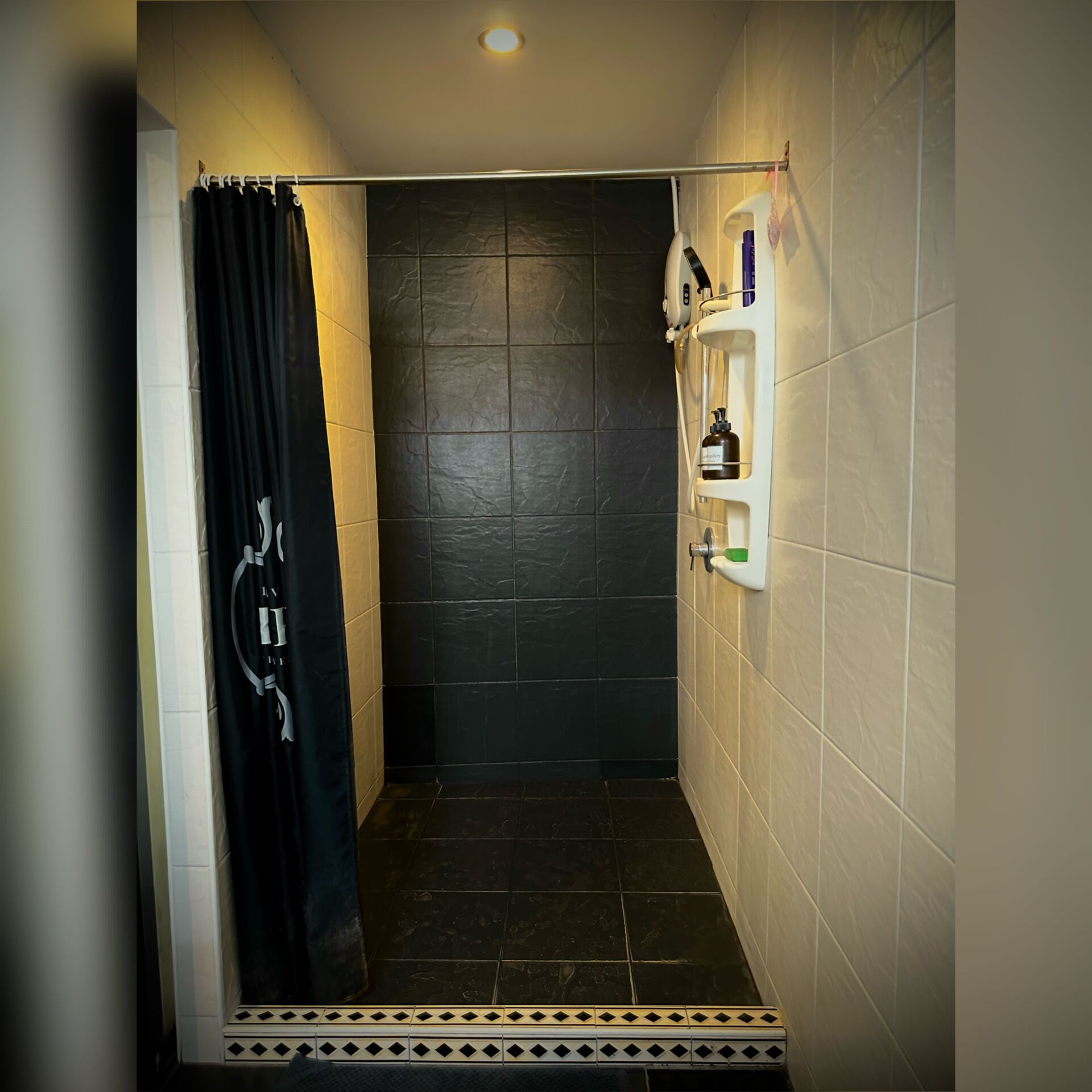 Espaço de duche estreito com azulejos escuros, cortina de chuveiro preta e prateleira com produtos de higiene pessoal.