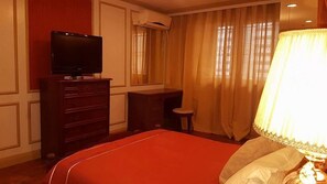 Quarto de hotel com cama, cômoda, televisão, escrivaninha, cadeira e abajur.