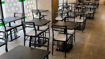 Interior de restaurante com várias mesas e cadeiras, janelas com padrão e luminárias pendentes.