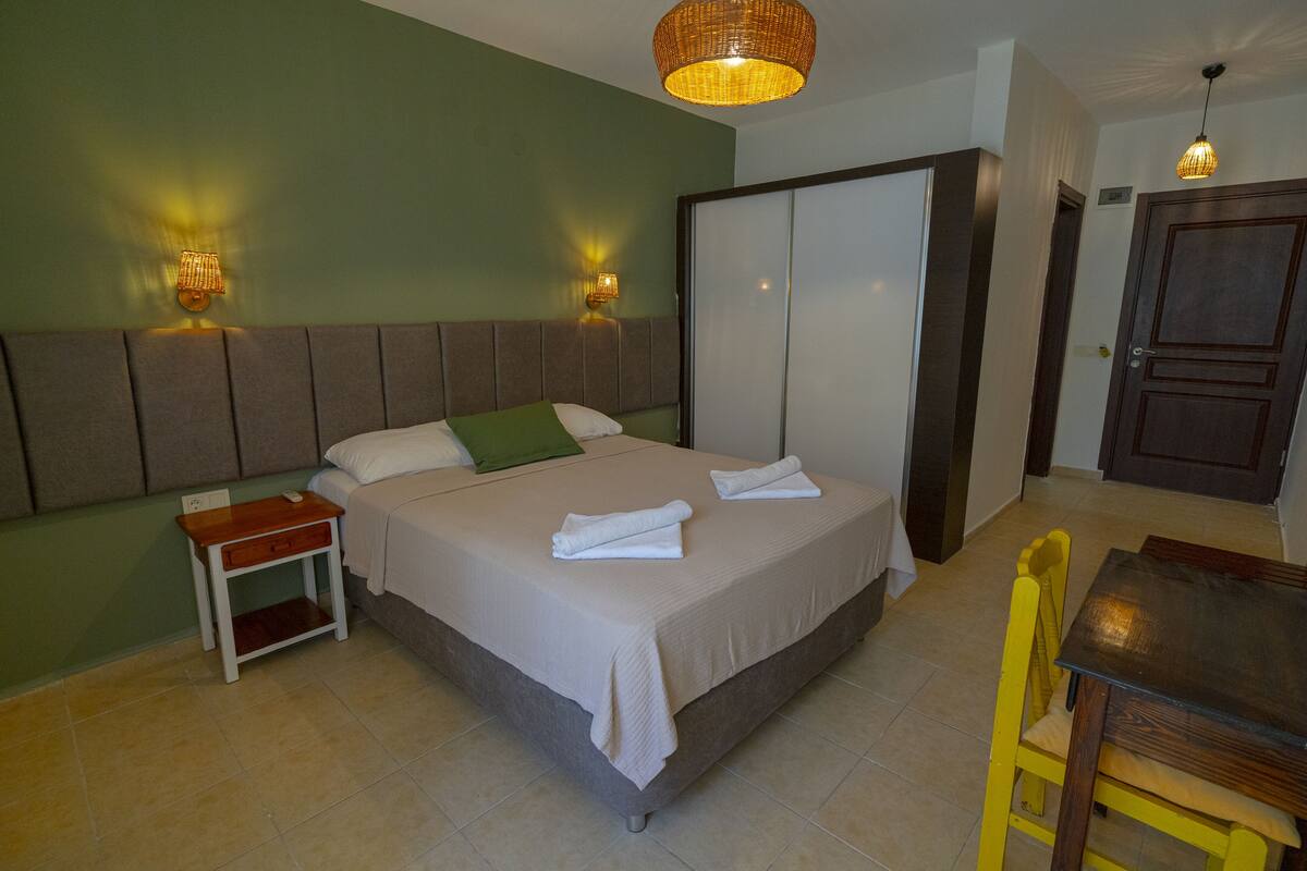 Cama arrumada com travesseiros e toalhas dobradas, mesa de cabeceira, cadeira e guarda-roupa.