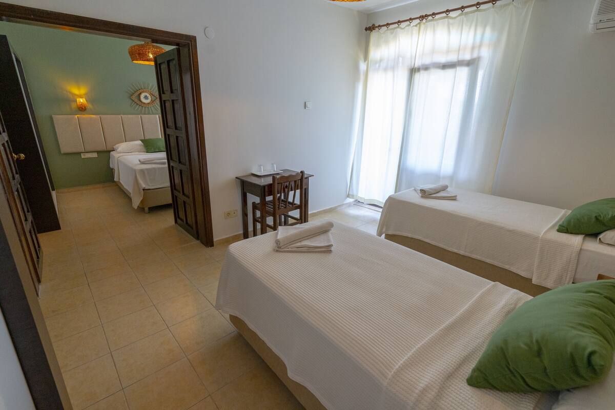 Quarto de hotel com duas camas, uma mesa pequena, uma cadeira e uma janela com cortinas.