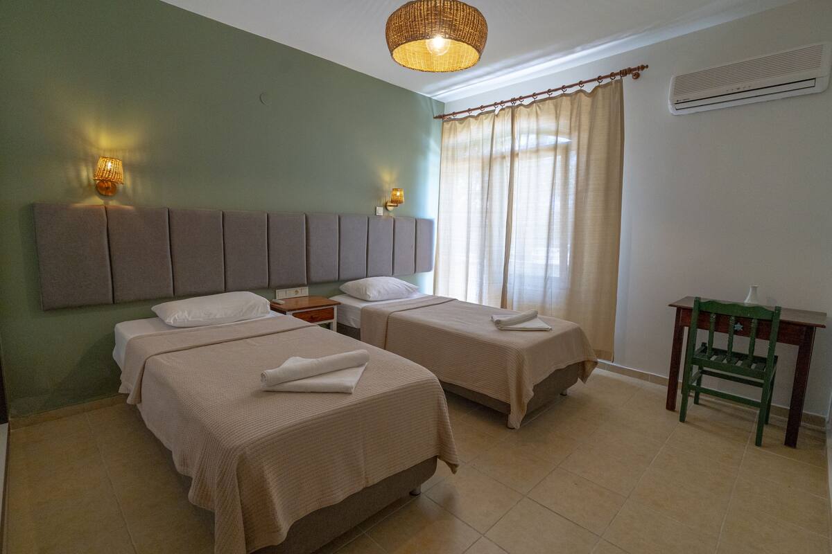 Quarto de hotel com duas camas, uma mesinha de cabeceira, uma luminária, uma cadeira e uma janela com cortinas.