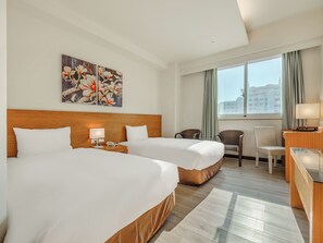Quarto de hotel com duas camas, uma escrivaninha, uma cadeira, uma janela com vista para edifícios e um quadro na parede.