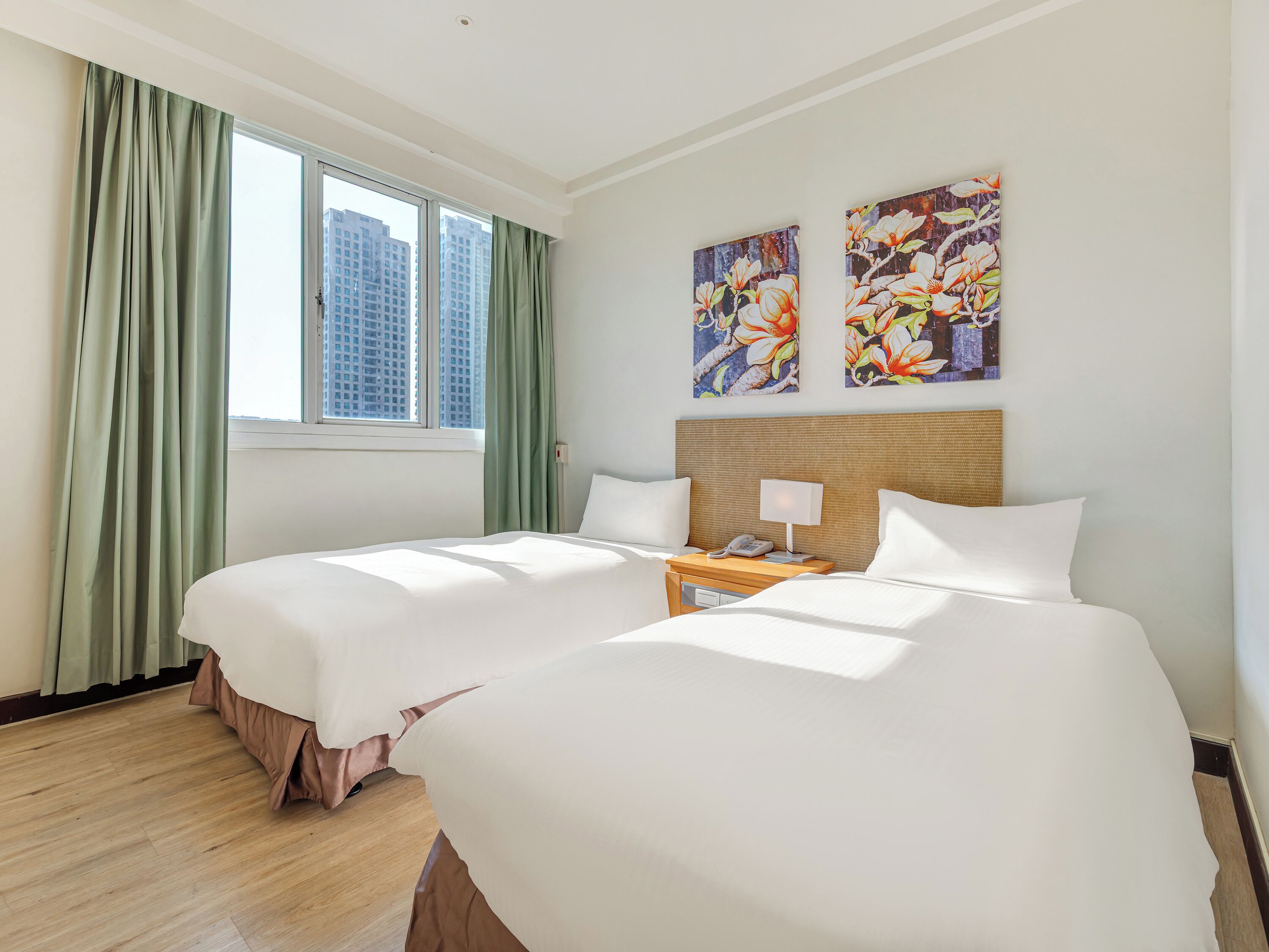 Quarto de hotel com duas camas, uma mesa de cabeceira com abajur e duas obras de arte floral na parede.