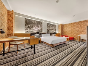 Quarto de hotel com cama, escrivaninha, cadeira e uma grande obra de arte abstrata na parede.