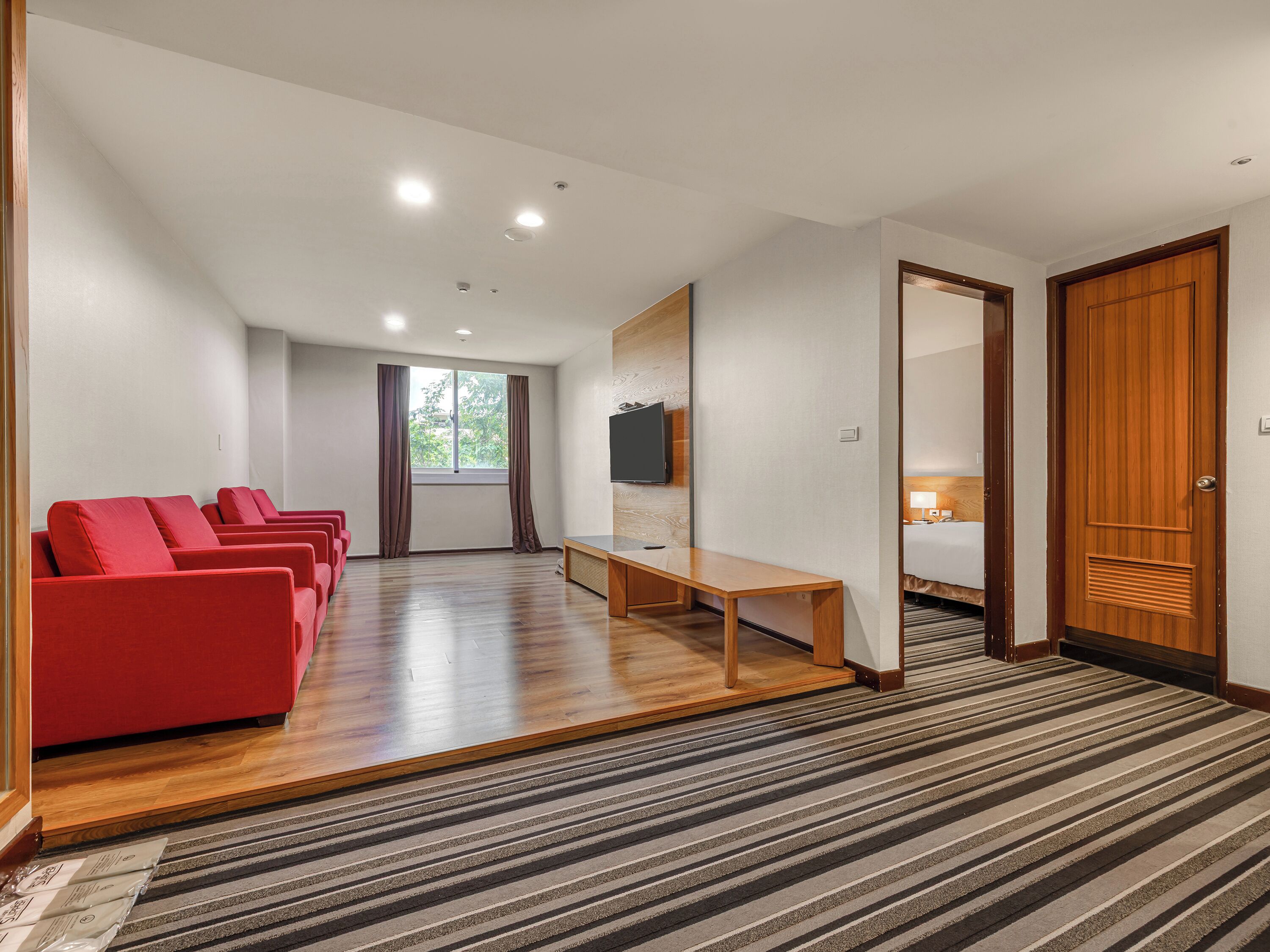 Quarto de hotel com piso de madeira, televisão fixada na parede, cama, escrivaninha e área de estar com cadeiras vermelhas.