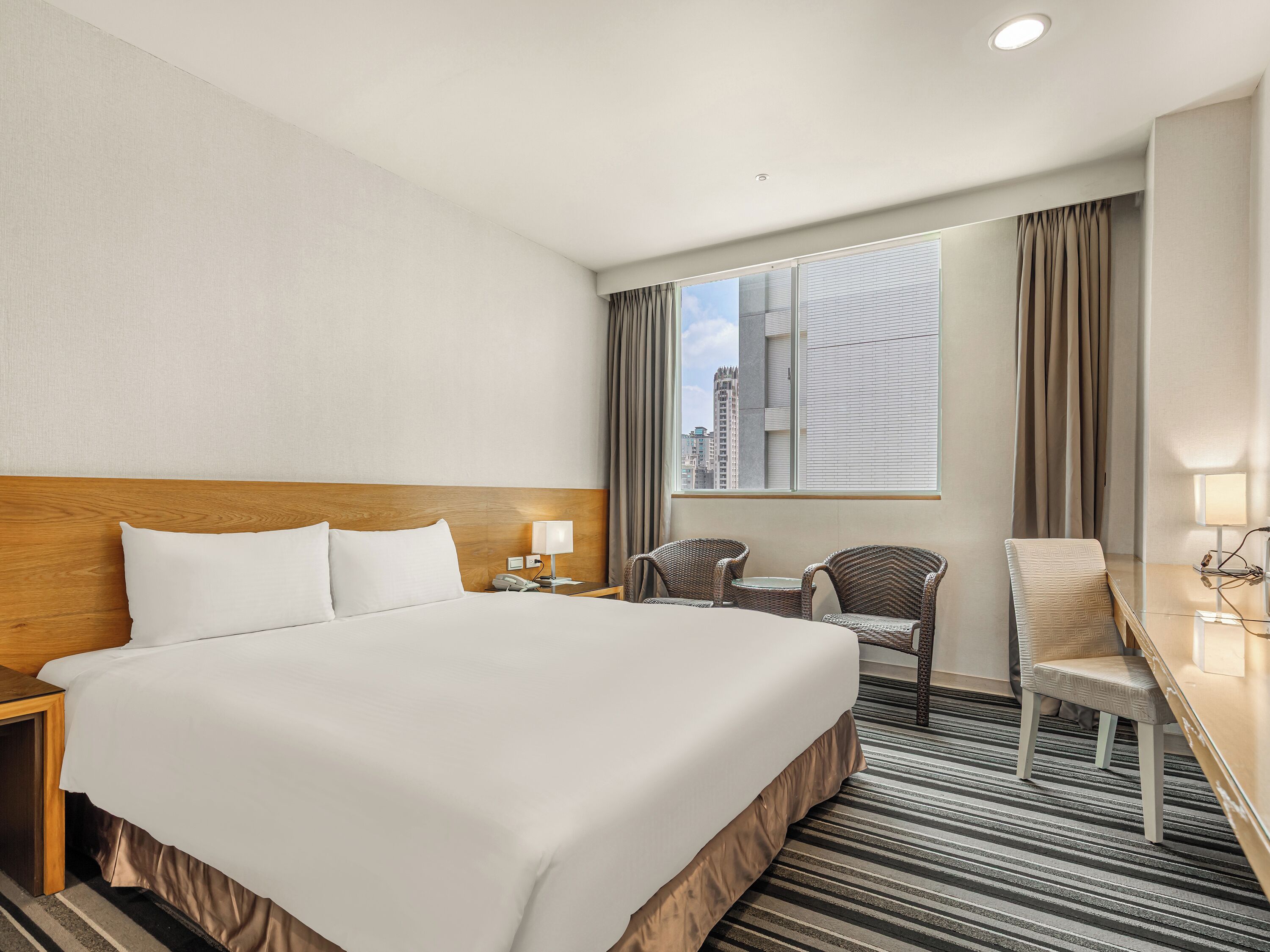Quarto de hotel com uma cama grande, uma mesa com cadeira, janela com cortinas e vista para a cidade.