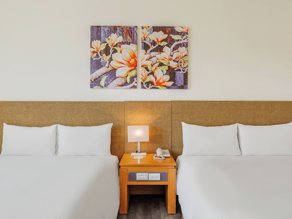 Quarto de hotel com duas camas, uma mesa de cabeceira com abajur e um quadro de flores na parede.