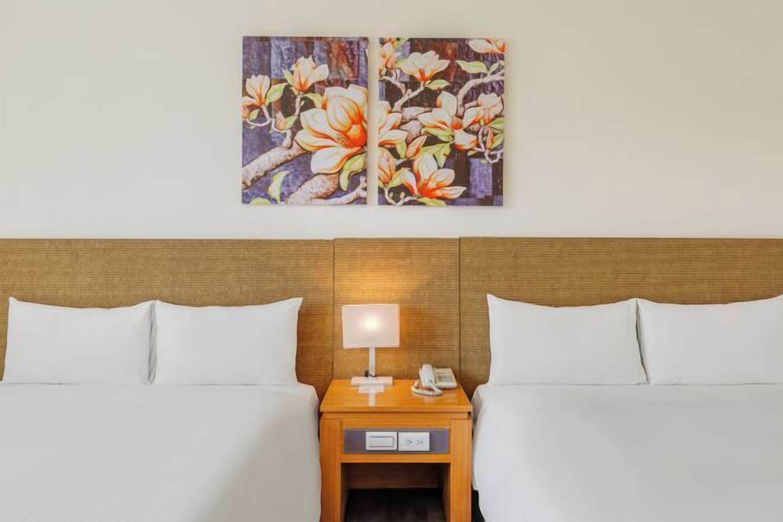 Quarto de hotel com duas camas, uma mesinha de cabeceira com abajur e uma obra de arte floral na parede.
