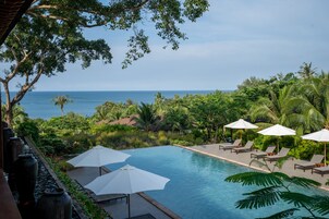 Área de piscina com espreguiçadeiras e guarda-sóis, com vista para uma paisagem tropical e o oceano.