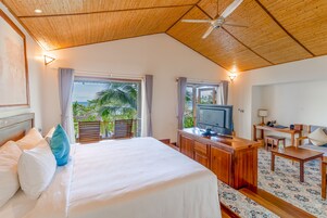 Quarto de hotel com cama, escrivaninha, televisão e varanda com vista para palmeiras.