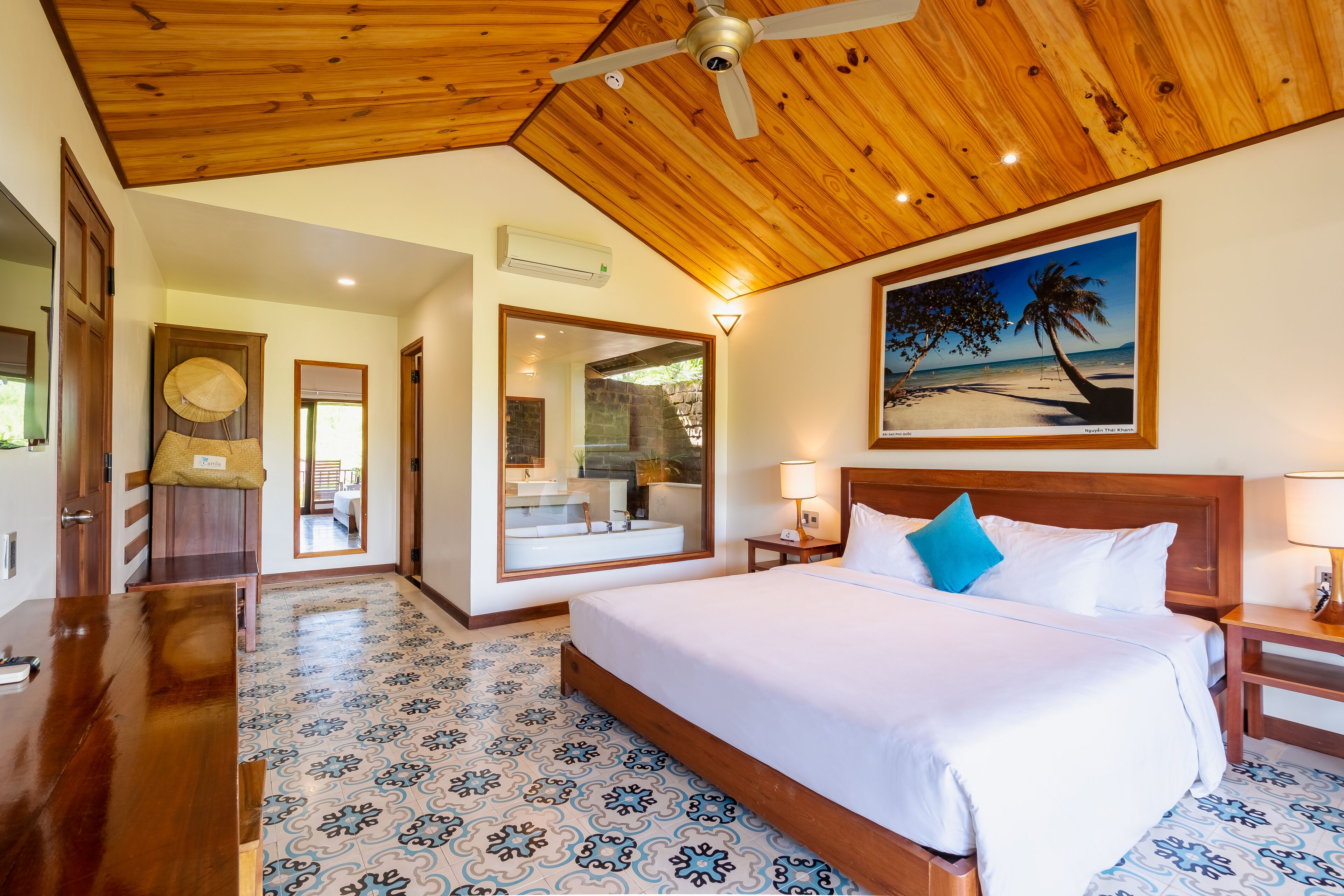 Quarto de hotel com teto de madeira, uma cama grande com roupa de cama branca e azul, um quadro de uma praia e um banheiro visível através de uma porta de vidro.