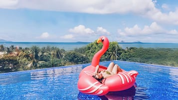 Uma pessoa relaxando em um flutuador de flamingos rosa em uma piscina infinita azul cristalina, com vista para o oceano e montanhas.